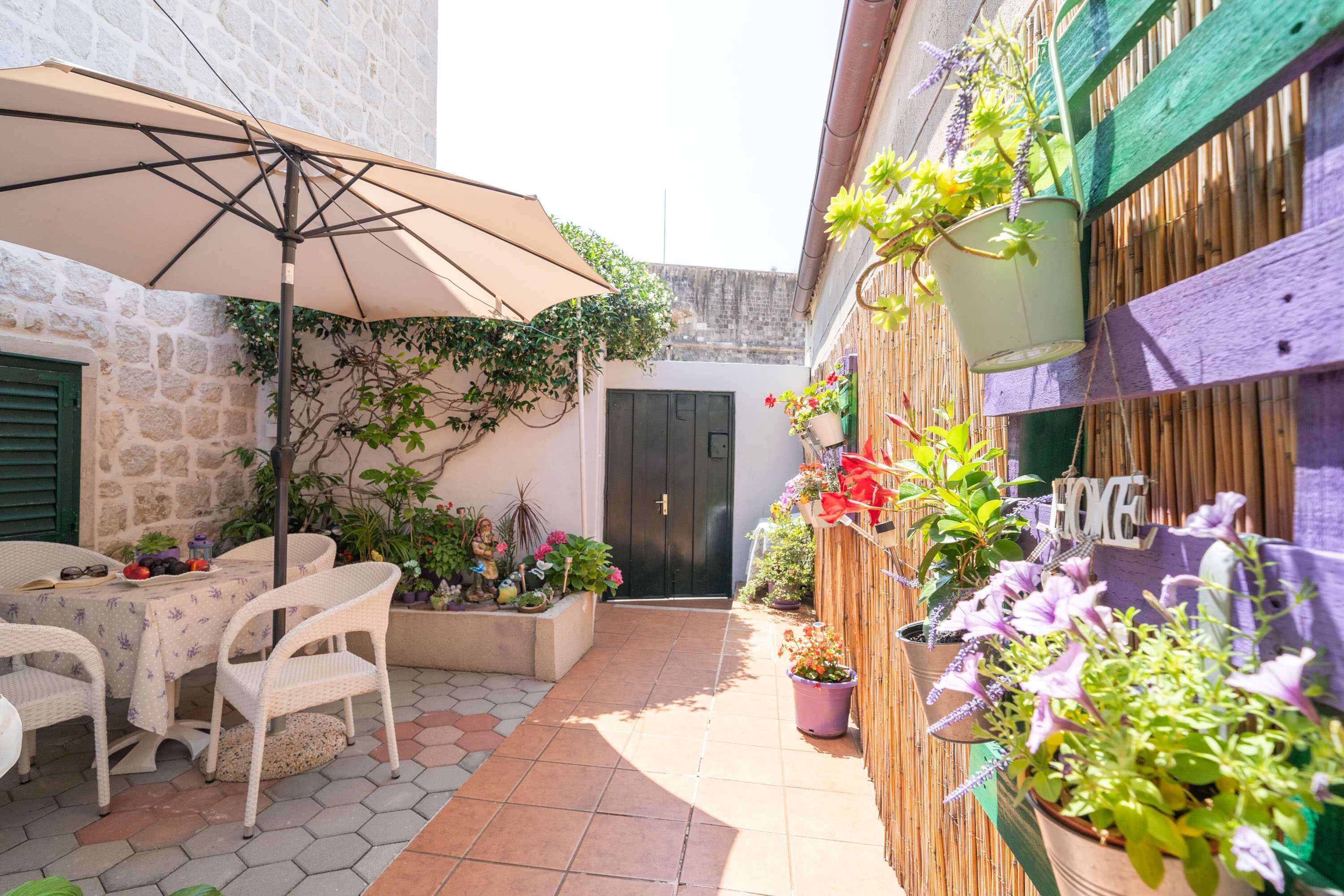 Um pátio com mesa e cadeiras sob um guarda-sol, rodeado por plantas em vasos e um cercado de madeira.