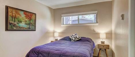 Quarto com cama, janela, mesa de cabeceira, luminária e um quadro na parede.