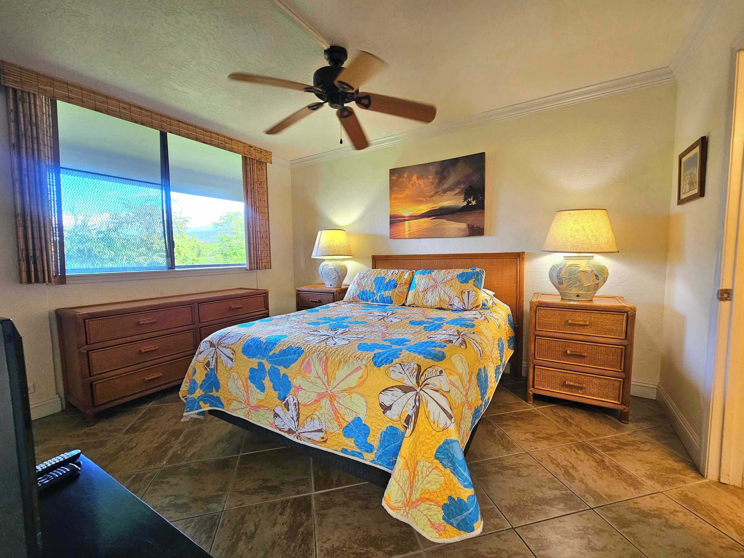 Um quarto com uma cama, duas mesas de cabeceira, um ventilador de teto, uma janela com cortinas, um quadro na parede e uma televisão sobre um móvel.