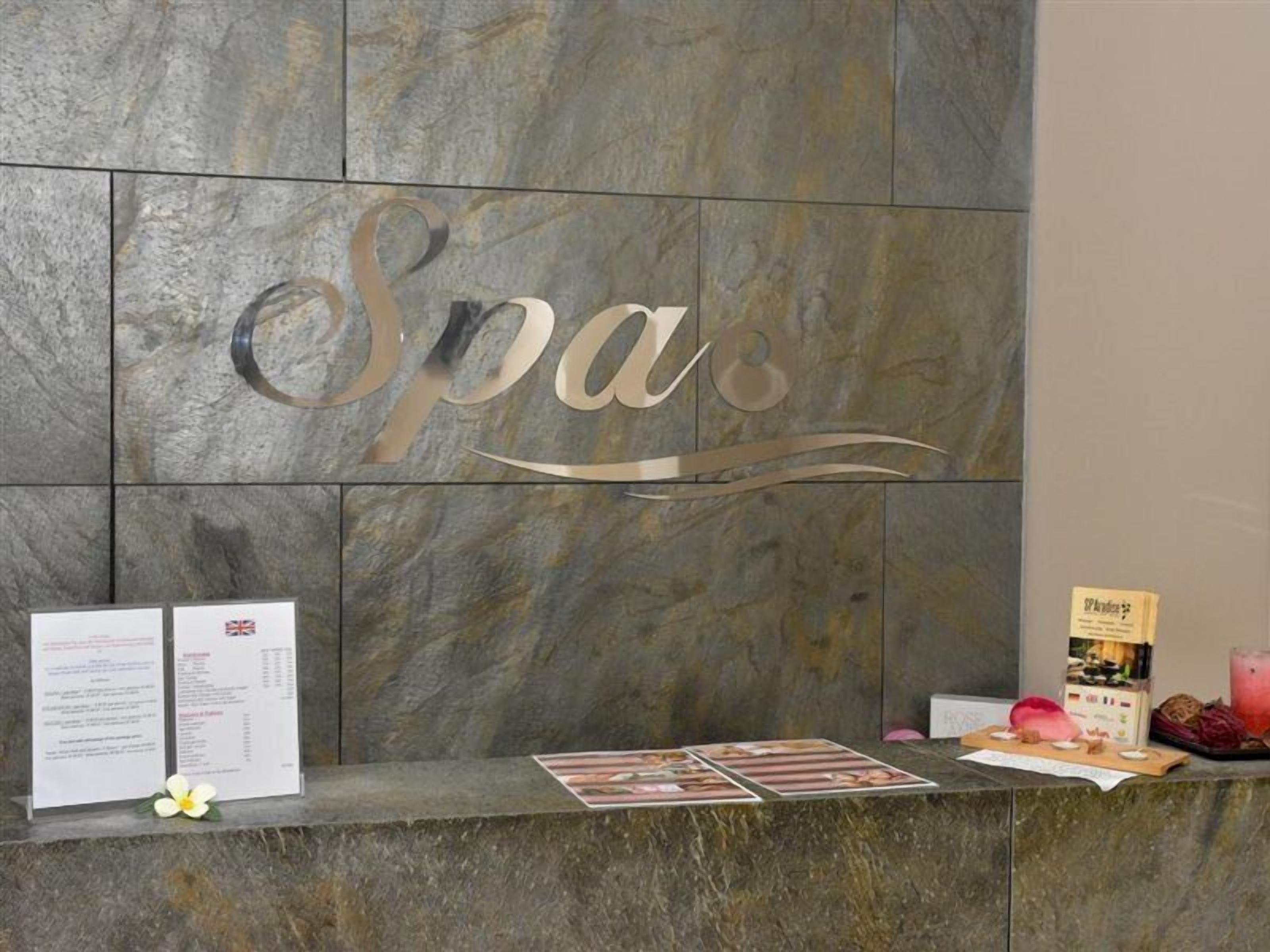 Uma receção com parede de pedra ao fundo, exibindo a palavra "Spa" numa fonte cursiva. Existem folhetos informativos e uma flor sobre a mesa.