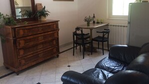 Uma sala de estar com um aparador de madeira, uma mesa de jantar com cadeiras, um sofá de couro preto, uma janela com uma planta e um quadro na parede.