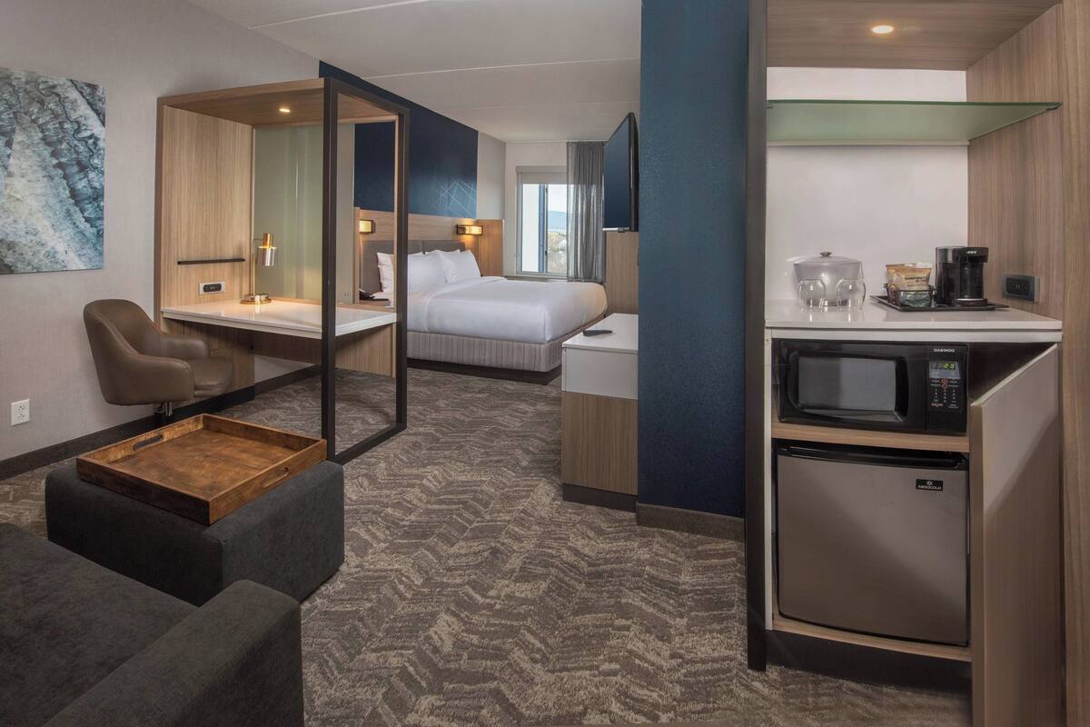 Quarto de hotel com cama, escrivaninha, cadeira, pequena cozinha e vista para o exterior.