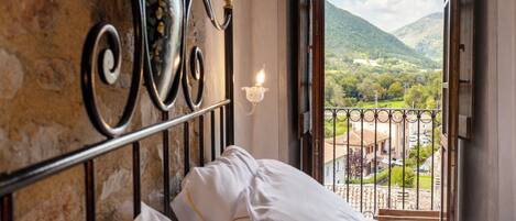Um quarto com cama, vista de uma paisagem montanhosa por uma porta aberta e uma varanda com guarda-corpo.