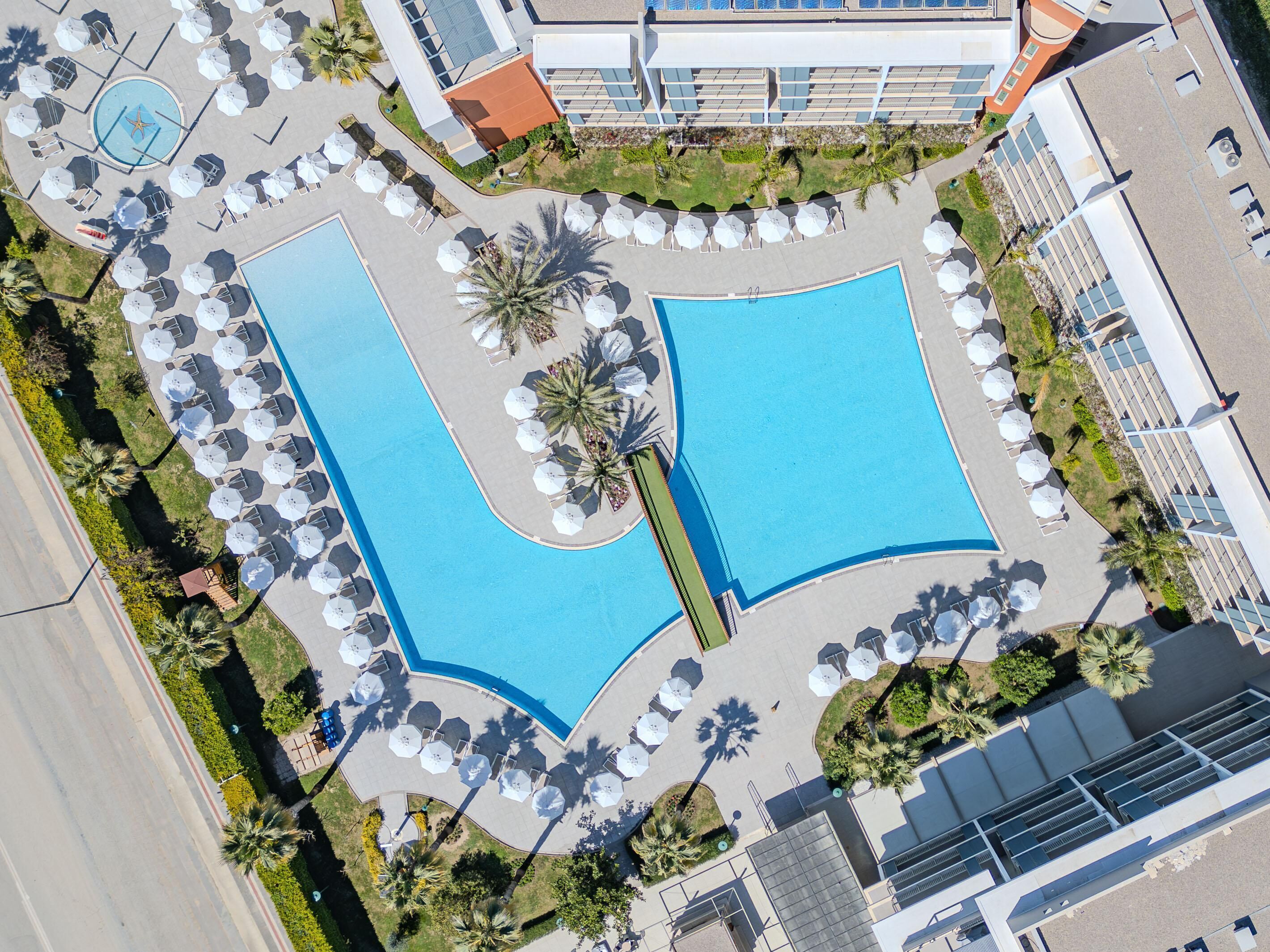 Vista aérea de um resort com duas piscinas grandes, cercado por guarda-sóis e palmeiras.