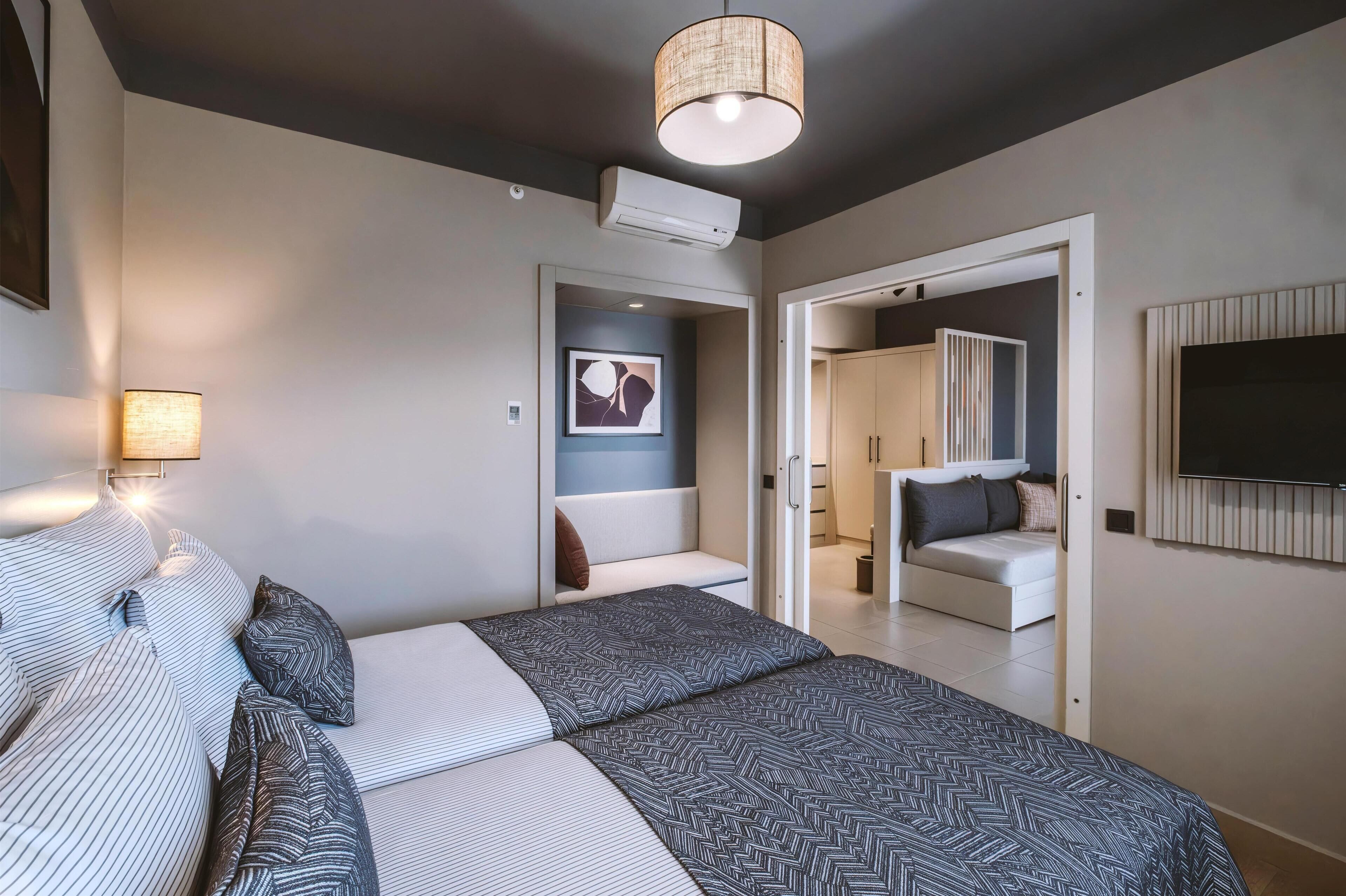 Quarto de hotel moderno com uma cama grande, área de estar, televisão e ar condicionado.