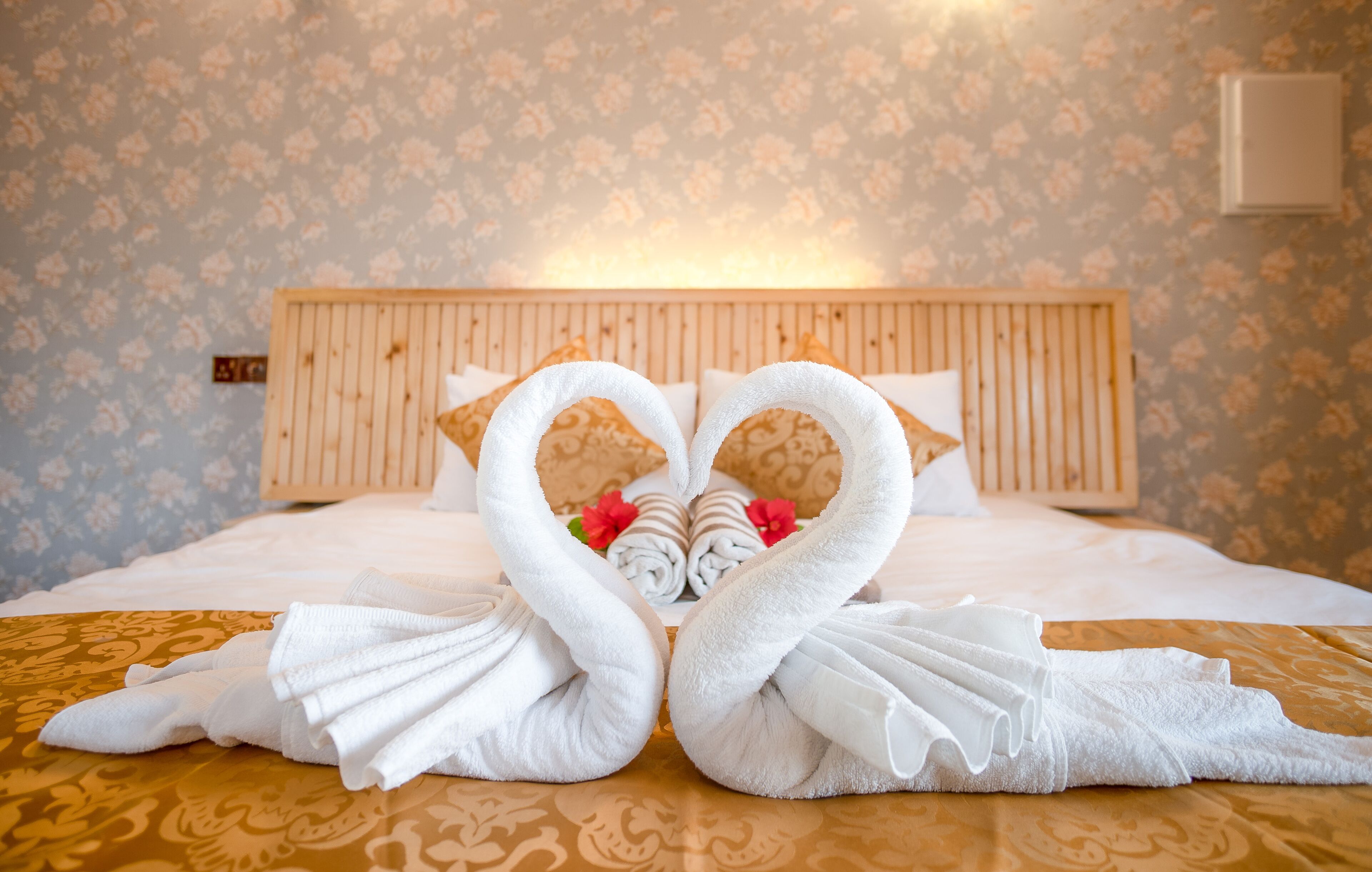 Uma cama com cabeceira de madeira, papel de parede com padrão floral e toalhas dispostas em formato de coração sobre a cama.