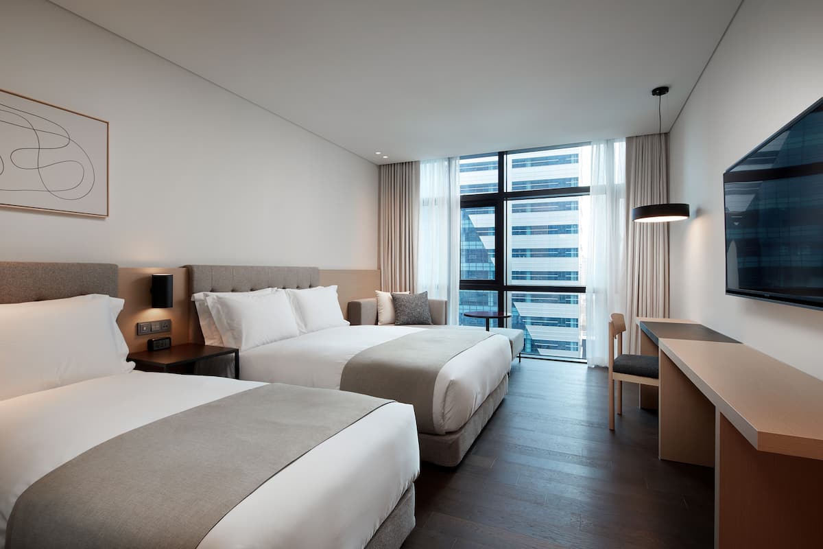 Quarto de hotel com duas camas, uma escrivaninha, uma cadeira, uma TV e uma janela grande com vista para a cidade.