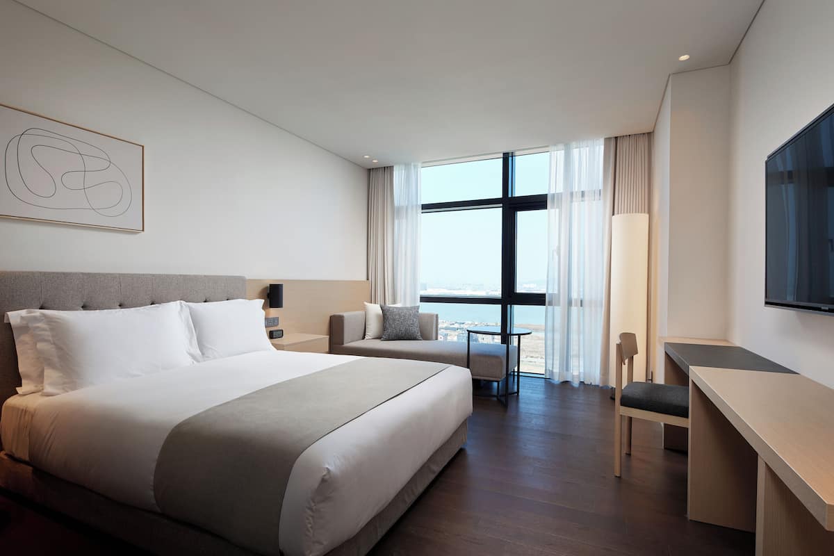 Quarto de hotel moderno com uma cama grande, uma escrivaninha e vista para o mar através de amplas janelas.