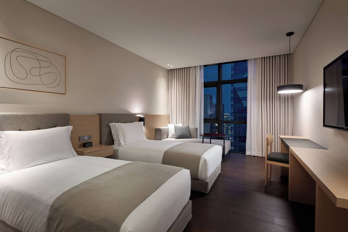 Quarto de hotel com duas camas, uma escrivaninha, uma cadeira, televisão e janela com cortinas.