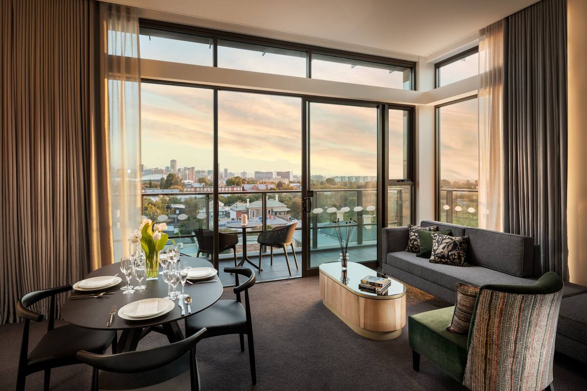 Quarto de hotel moderno com área de refeições, sofá e uma grande janela com vista para a cidade.