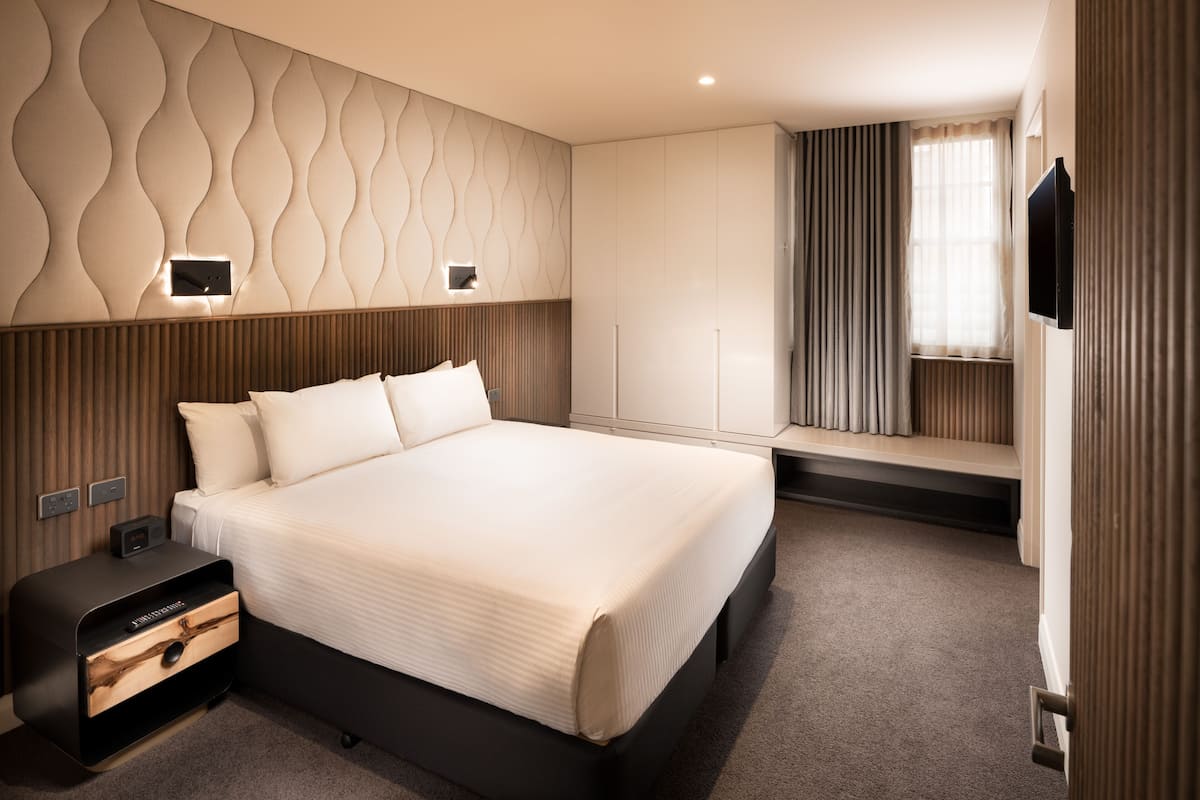 Quarto de hotel moderno com uma cama grande, mesa de cabeceira de madeira, um banco embutido e uma TV de tela plana na parede.