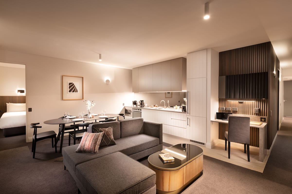 Quarto de hotel moderno com sofá, mesa de centro e área de jantar com cozinha ao fundo.