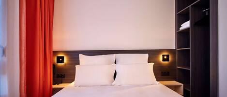 Quarto de hotel com cama bem arrumada, duas mesas de cabeceira e um guarda-roupa com cabides.