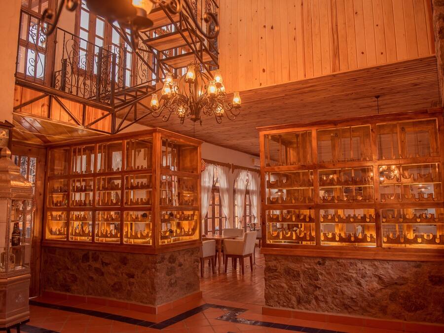 Um quarto iluminado com luz quente, paredes de madeira e um candelabro, com uma mesa central e cadeiras, e prateleiras com armários de vidro exibindo diversos objetos.