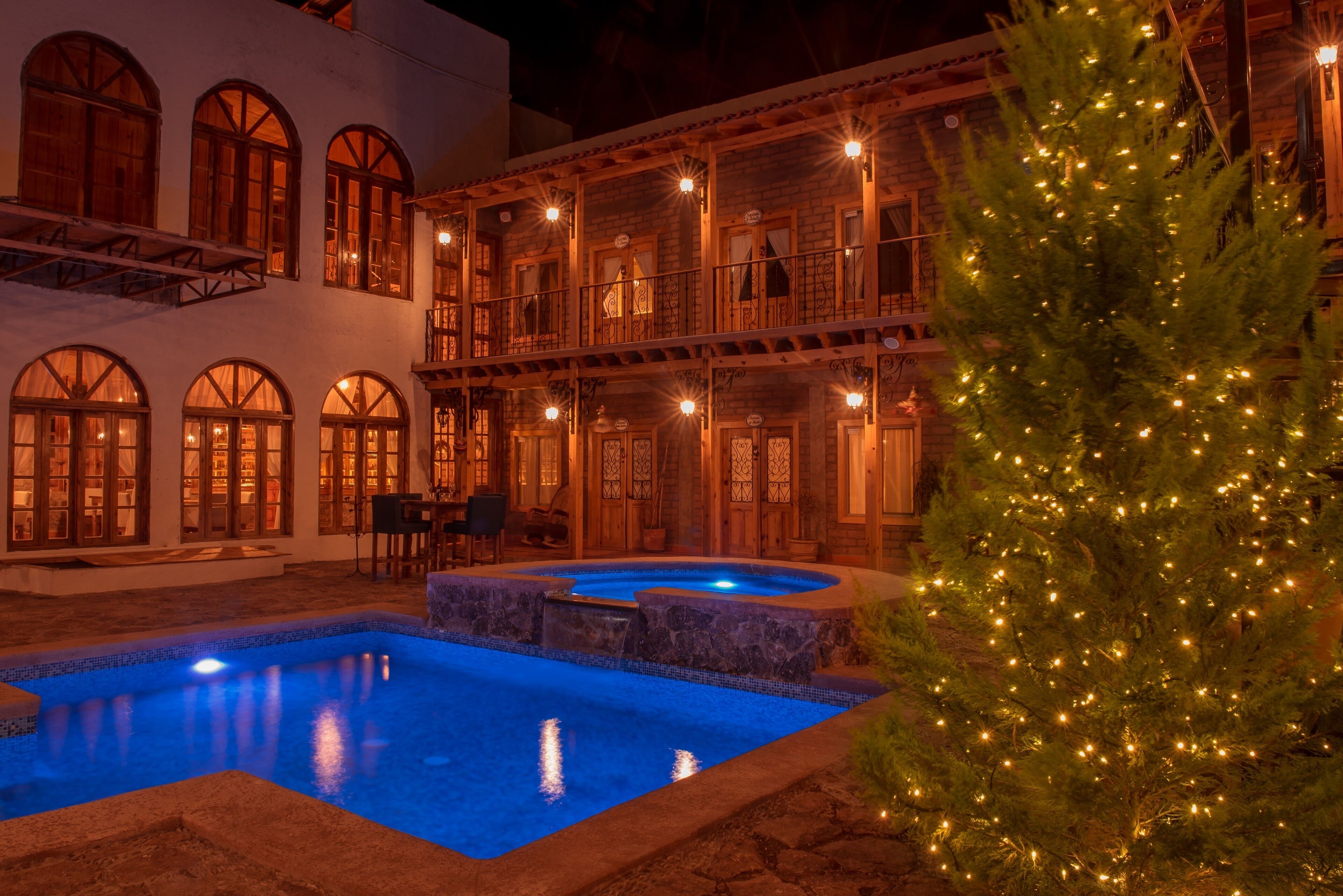 Uma área de piscina com banheira de hidromassagem, uma árvore de Natal e um edifício com janelas em arco e varandas.