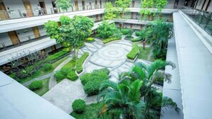 Pátio com vários níveis e vegetação exuberante, incluindo palmeiras e arbustos diversos, rodeado por um edifício moderno com varandas.