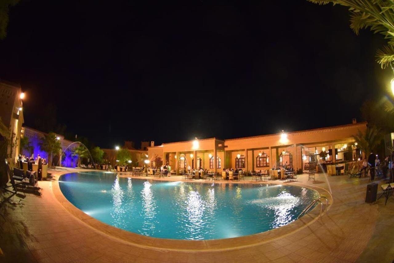 Uma área de piscina à noite, com espaço de estar coberto e um edifício com várias janelas e portas.