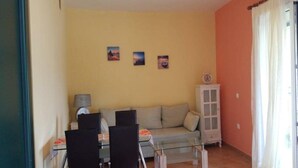 Uma sala de estar com sofá, mesa de vidro e cadeiras. Há três quadros pendurados na parede.