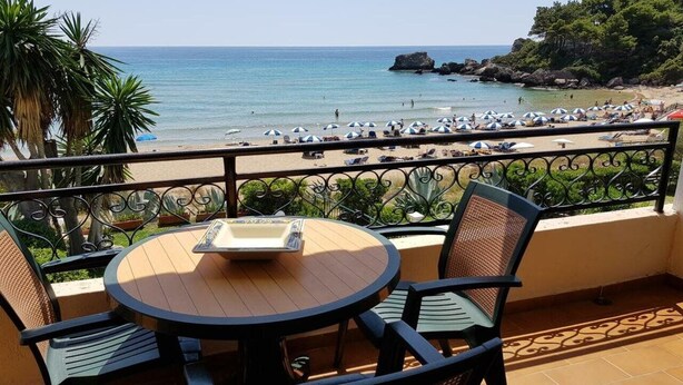 Varanda com mesa e cadeiras redondas, vista para uma praia com guarda-sóis e pessoas.