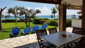 Um pátio externo com mesa e cadeiras, com vista para uma área gramada com árvores e o mar.