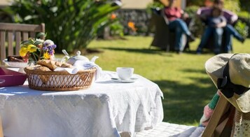 Uma mesa com uma cesta de pão, um bule e xícaras, disposta ao ar livre, com gramado e um banco de madeira ao fundo.
