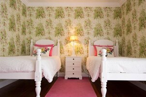 Quarto com duas camas brancas, uma mesinha de cabeceira e papel de parede floral.
