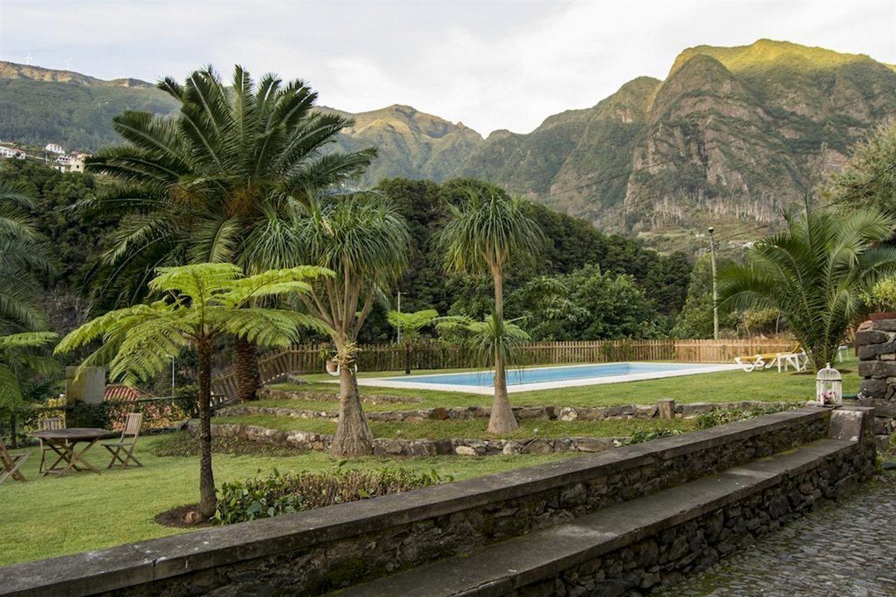 Uma piscina rodeada por palmeiras e uma parede de pedra, com montanhas ao fundo.