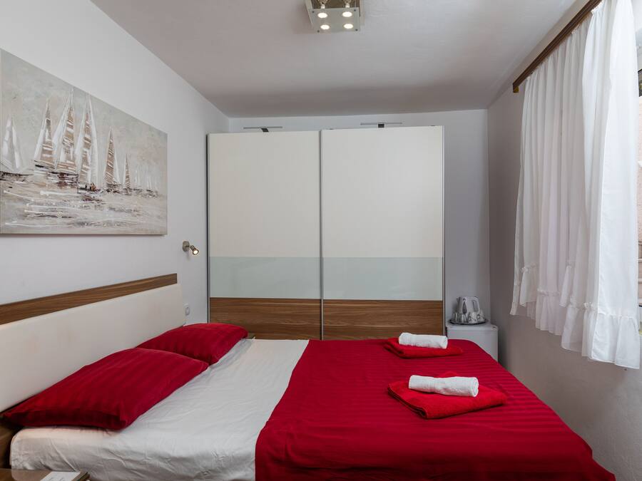 Quarto de hotel com cama, roupa de cama vermelha, um quadro na parede, um guarda-roupa e uma janela com cortinas.