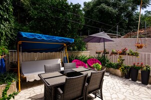 Um pátio externo com mesa e cadeiras de vime, um toldo azul e plantas em vasos.