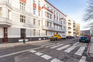 Uma rua com um caminhão amarelo estacionado na lateral, edifícios com várias janelas e varandas, e um céu claro.
