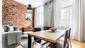Uma sala de estar moderna com parede de tijolos, sofá cinza, mesa de jantar de madeira e uma janela grande com cortinas.