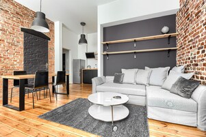 Uma sala de estar moderna com parede de tijolos, um sofá cinza, uma mesa de centro redonda e uma área de jantar com mesa e cadeiras de madeira.