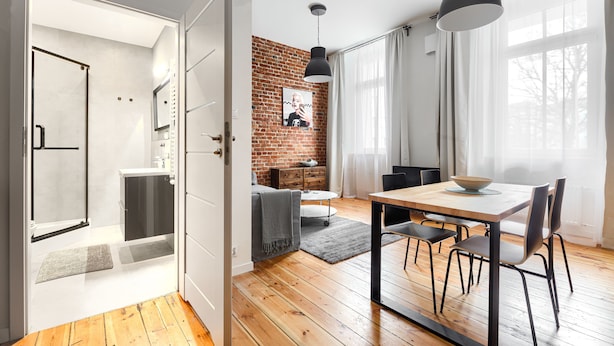 Interior moderno com piso de madeira, área de refeições com mesa e cadeiras, parede decorativa de tijolos e uma janela grande com cortinas.