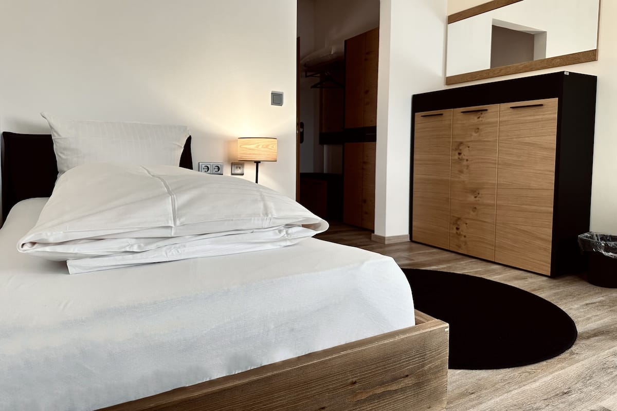 Cama bem arrumada com lençóis brancos, luminária de cabeceira, guarda-roupa de madeira e tapete preto sobre piso de madeira.