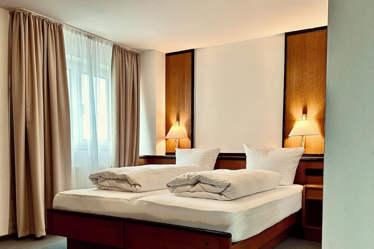 Quarto de hotel com uma cama grande, duas luminárias de cabeceira, uma luz de teto e uma janela com cortinas.