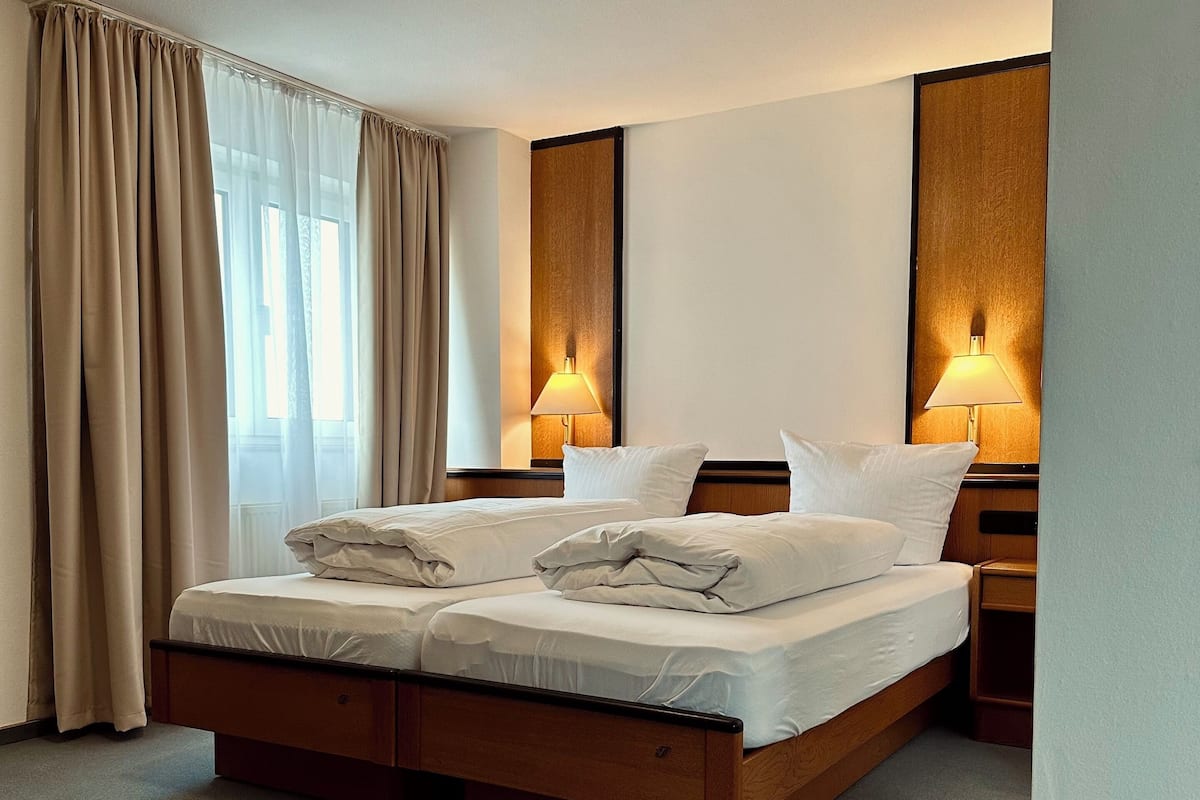 Quarto de hotel com cama de casal, duas mesas de cabeceira com abajures, janela com cortinas e luminária no teto.