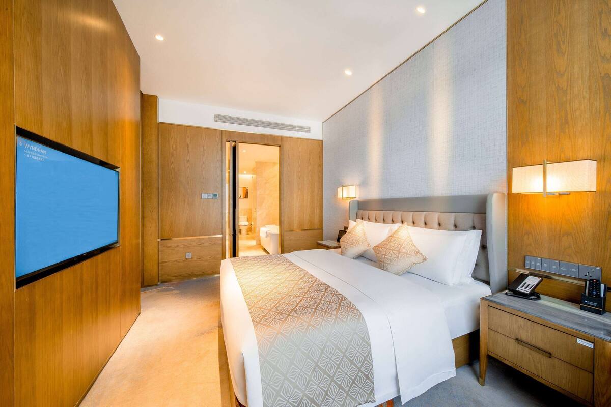 Quarto de hotel com uma cama grande, luminárias de cabeceira, mesa de cabeceira com telefone e televisão na parede.
