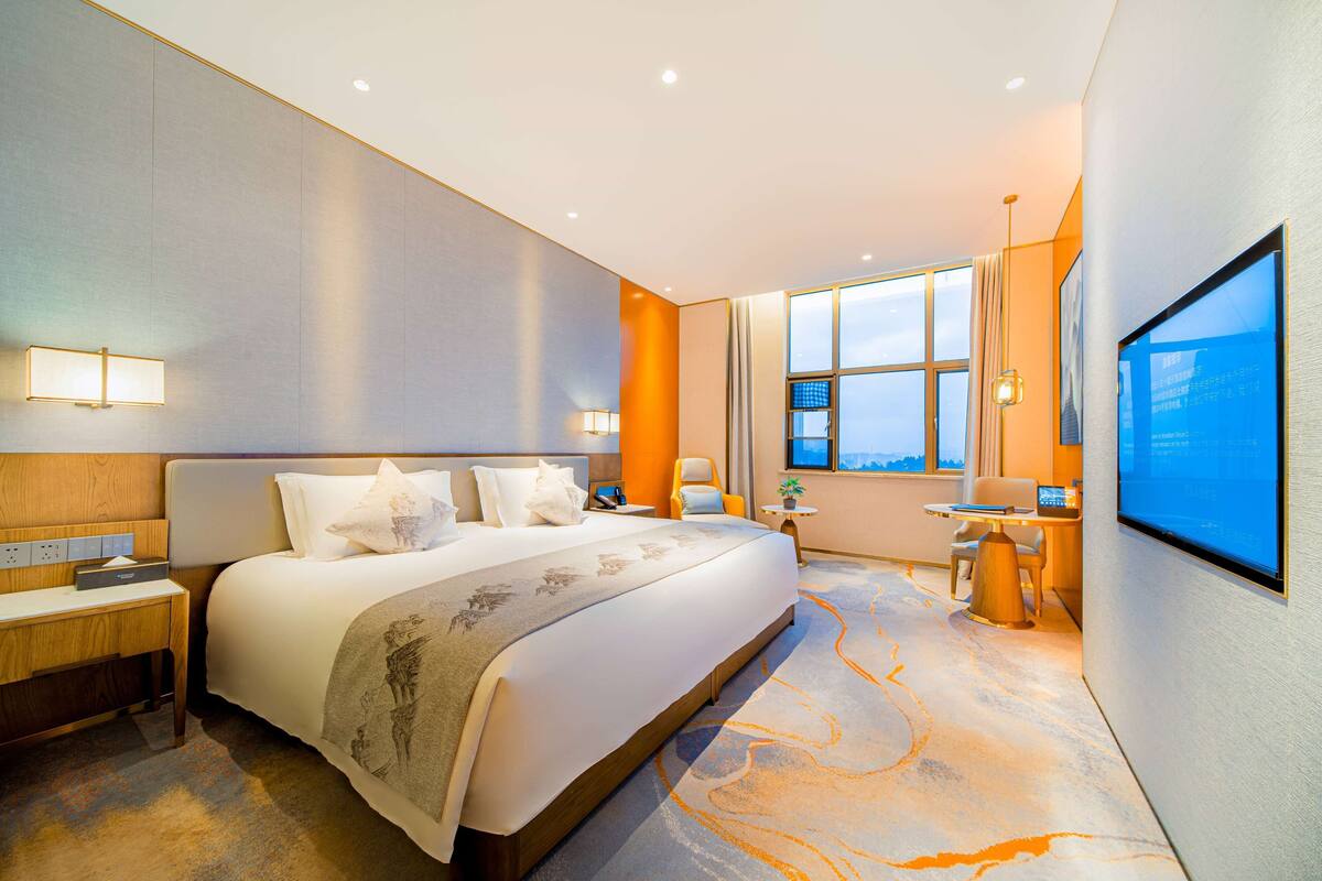Quarto de hotel moderno com uma cama grande, uma escrivaninha, uma cadeira, uma televisão e uma janela com vista para a cidade.