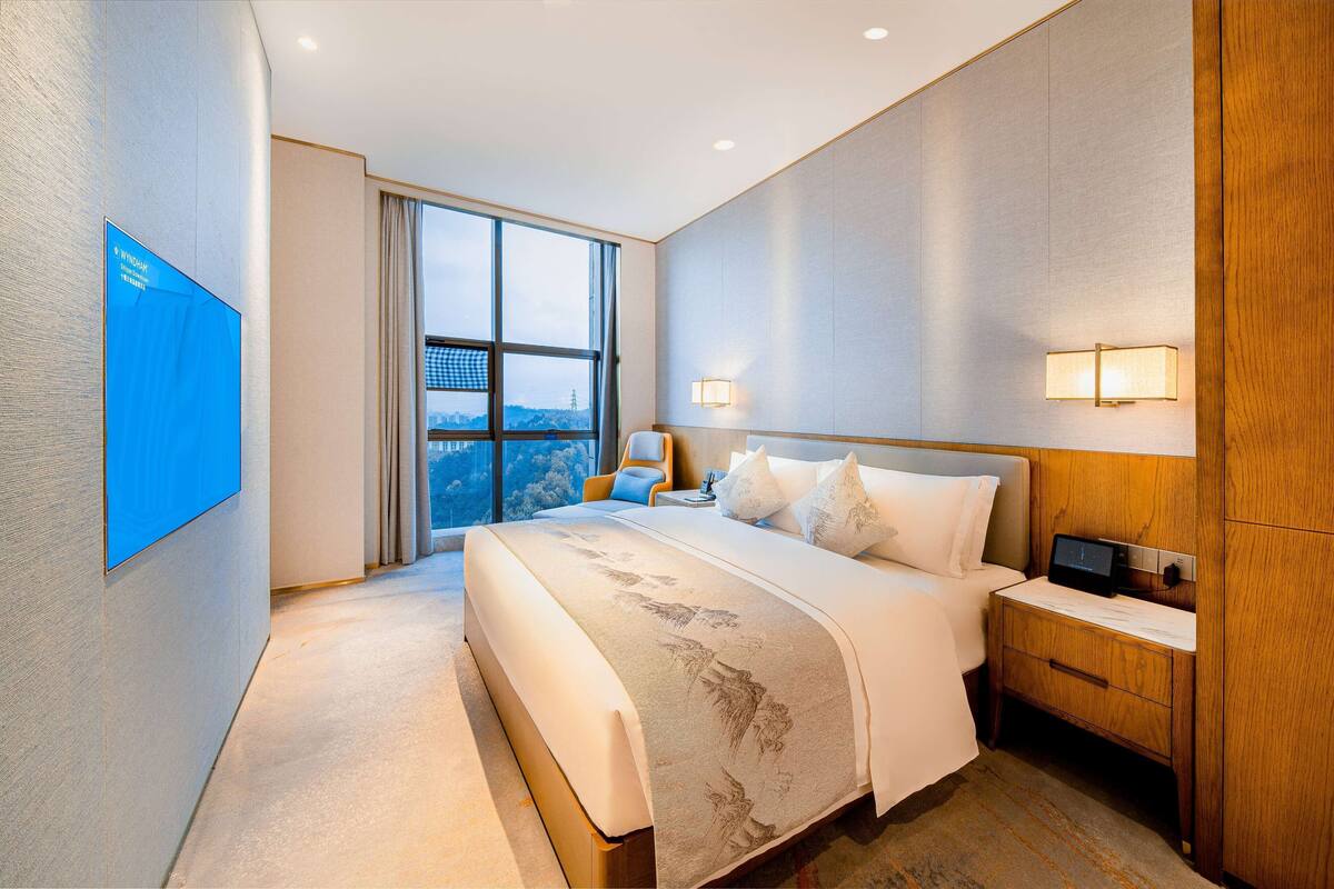 Quarto de hotel moderno com uma cama grande, televisão de tela plana, mesa de cabeceira com abajur e vista para a cidade pela janela.