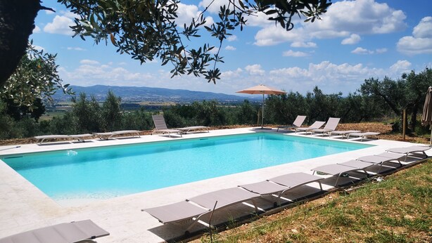 Piscina retangular com espreguiçadeiras e um guarda-sol, rodeada por vegetação e vista para montanhas ao longe.
