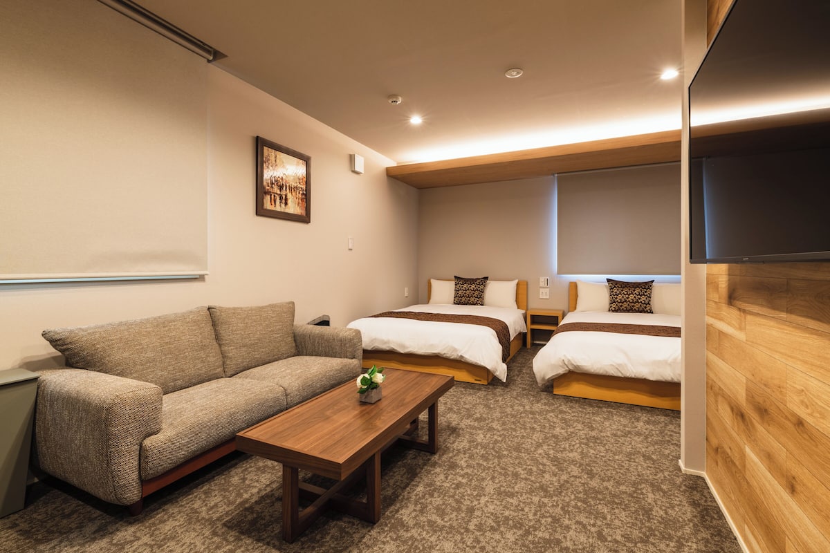 Quarto de hotel com sofá, duas camas de solteiro, mesa de centro de madeira e um quadro na parede.