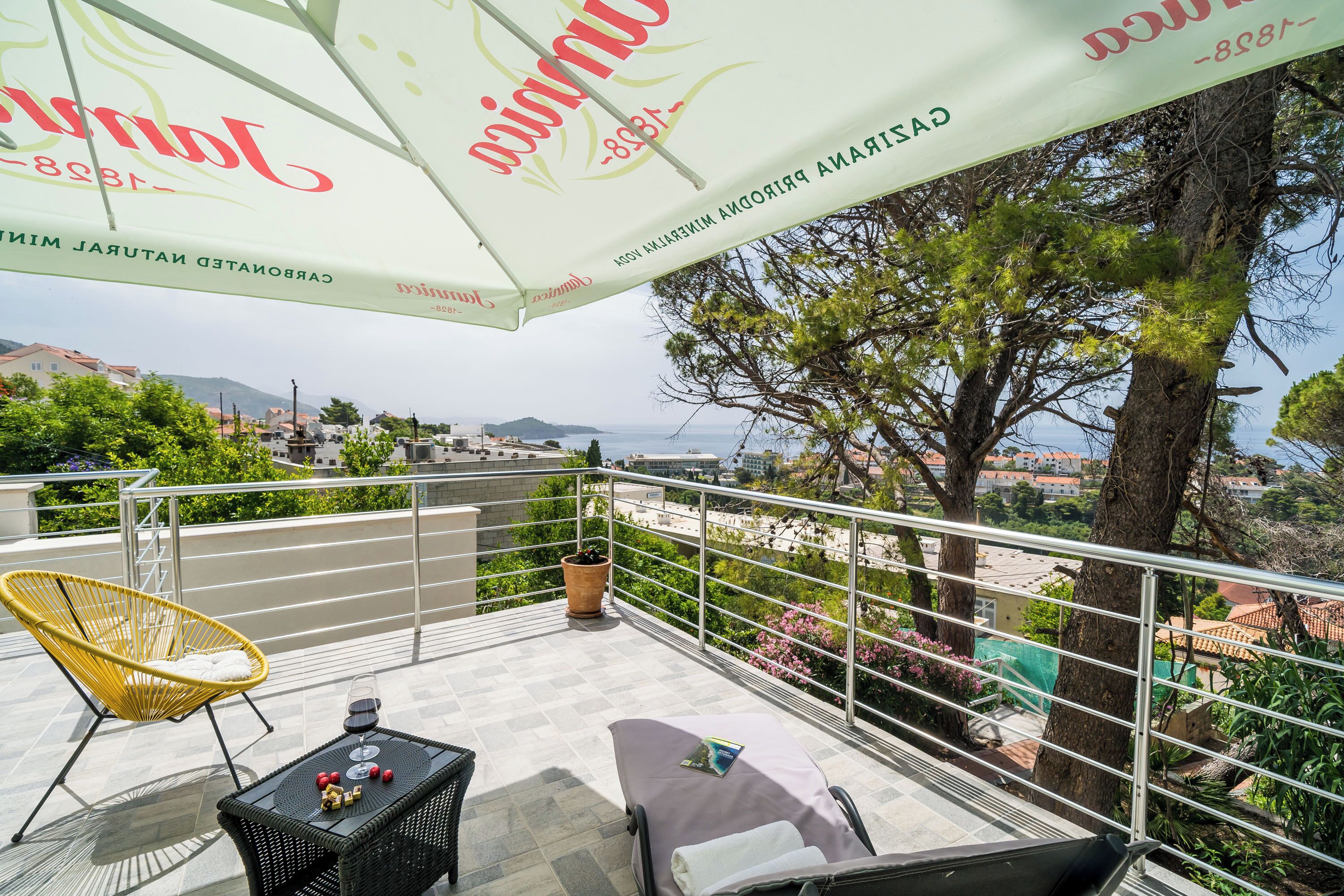 Uma varanda com uma cadeira amarela, uma mesa com um copo de vinho e vista para o mar e edifícios.