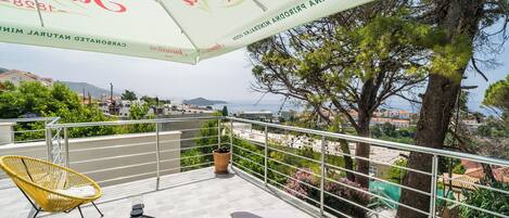 Uma varanda com uma cadeira amarela, uma mesa com um copo de vinho e vista para o mar e edifícios.