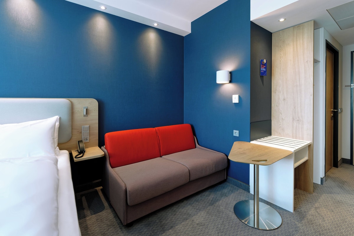Quarto de hotel com uma cama, um sofá vermelho e castanho, uma mesa pequena e uma luminária fixada na parede.