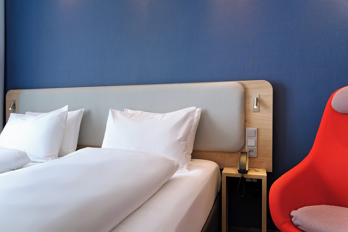 Quarto de hotel com uma cama, mesa de cabeceira e um telefone, além de uma cadeira vermelha.