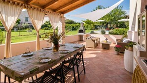 Um pátio externo com mesa de jantar posta para dois, rodeado por móveis de vime, uma lareira de pedra e vista para uma piscina e jardim.