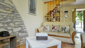 Uma sala de estar com uma parede de pedra, uma mesa branca, um sofá bege com almofadas florais e uma escadaria que leva a um andar superior.