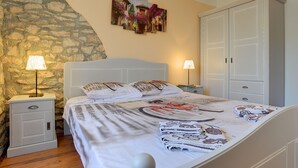 Quarto com parede de pedra, cama com cabeceira branca, duas mesas de cabeceira com luminárias e um guarda-roupa.