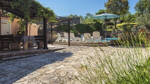 Uma área de varanda externa com piso de pedra, cadeiras de descanso, um pergolado com trepadeiras e uma piscina ao fundo.