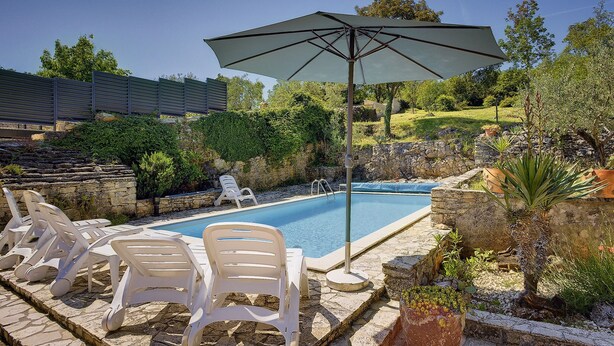 Piscina com espreguiçadeiras e um guarda-sol, cercada por vegetação e paredes de pedra.